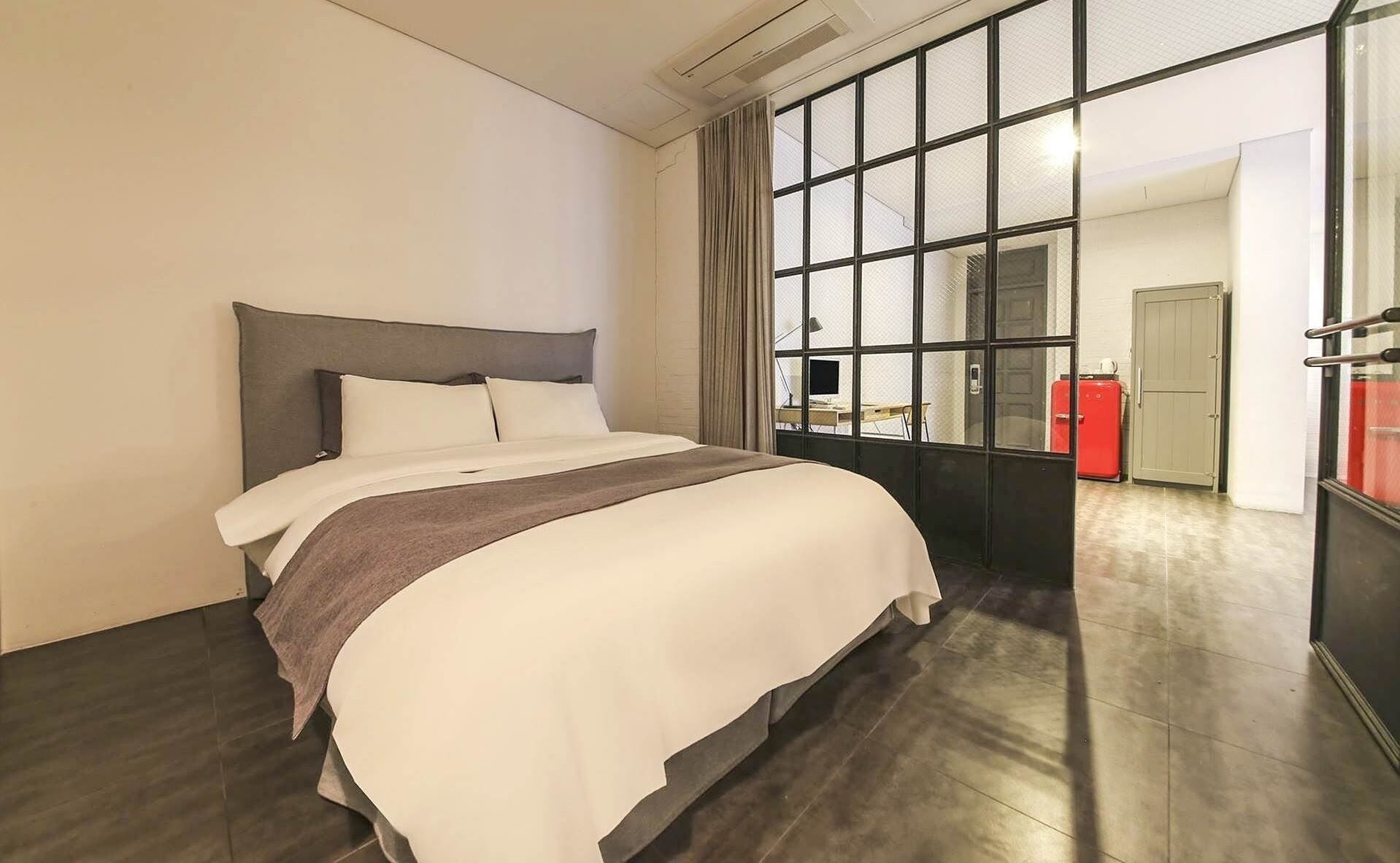 Quarto de hotel moderno com uma cama grande, uma janela com persianas, uma mesa com cadeira e um aparelho vermelho no canto.