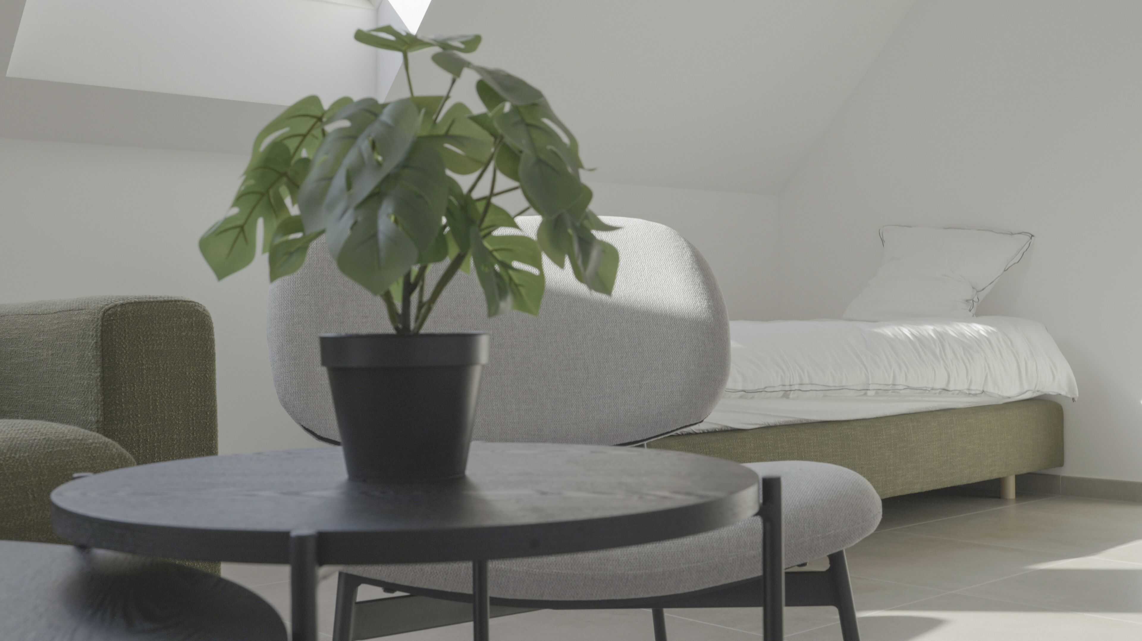 Quarto moderno e minimalista com uma mesa redonda, um vaso de planta, um sofá cinza e uma cama com roupa de cama branca.