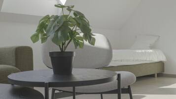 Quarto moderno e minimalista com uma mesa redonda, um vaso de planta, um sofá cinza e uma cama com roupa de cama branca.