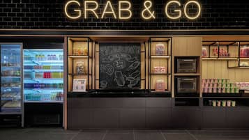 Uma lanchonete 'GRAB & GO' com um letreiro de neon, um quadro negro com o menu e prateleiras repletas de lanches e bebidas.