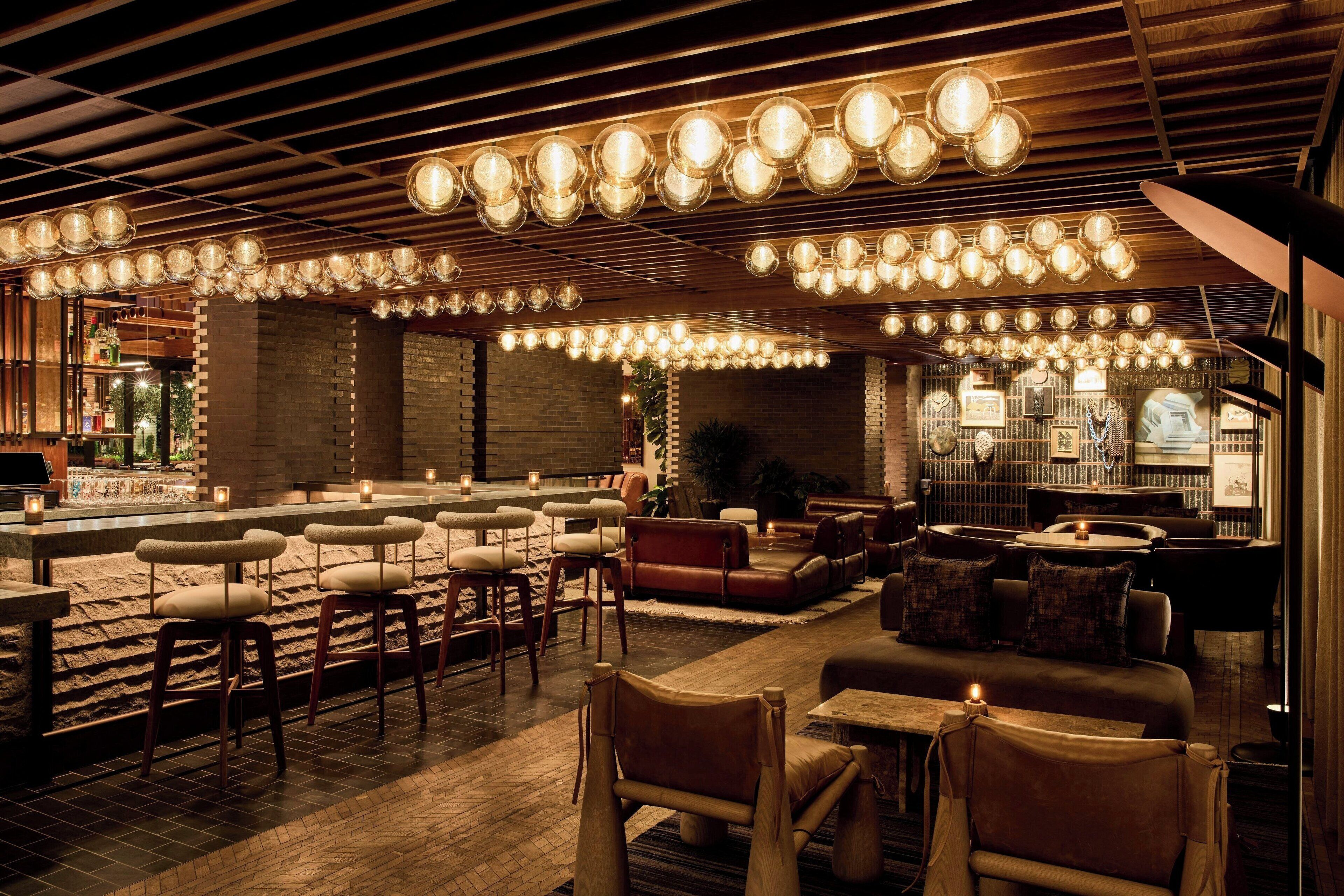Uma área de bar com diversas opções de assento, incluindo banquetas e sofás, e vários lustres pendentes.