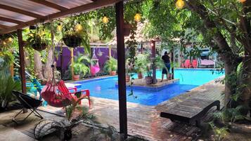 Piscina ao ar livre coberta com um deck de madeira, cercada por plantas em vasos e lâmpadas penduradas.