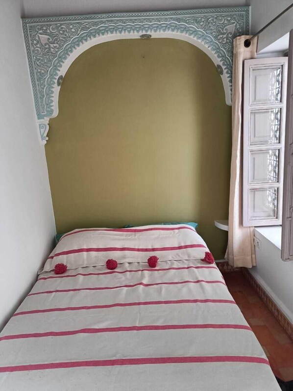 Uma cama com colcha listrada e detalhes vermelhos, uma cabeceira decorativa em arco e uma janela com cortinas.
