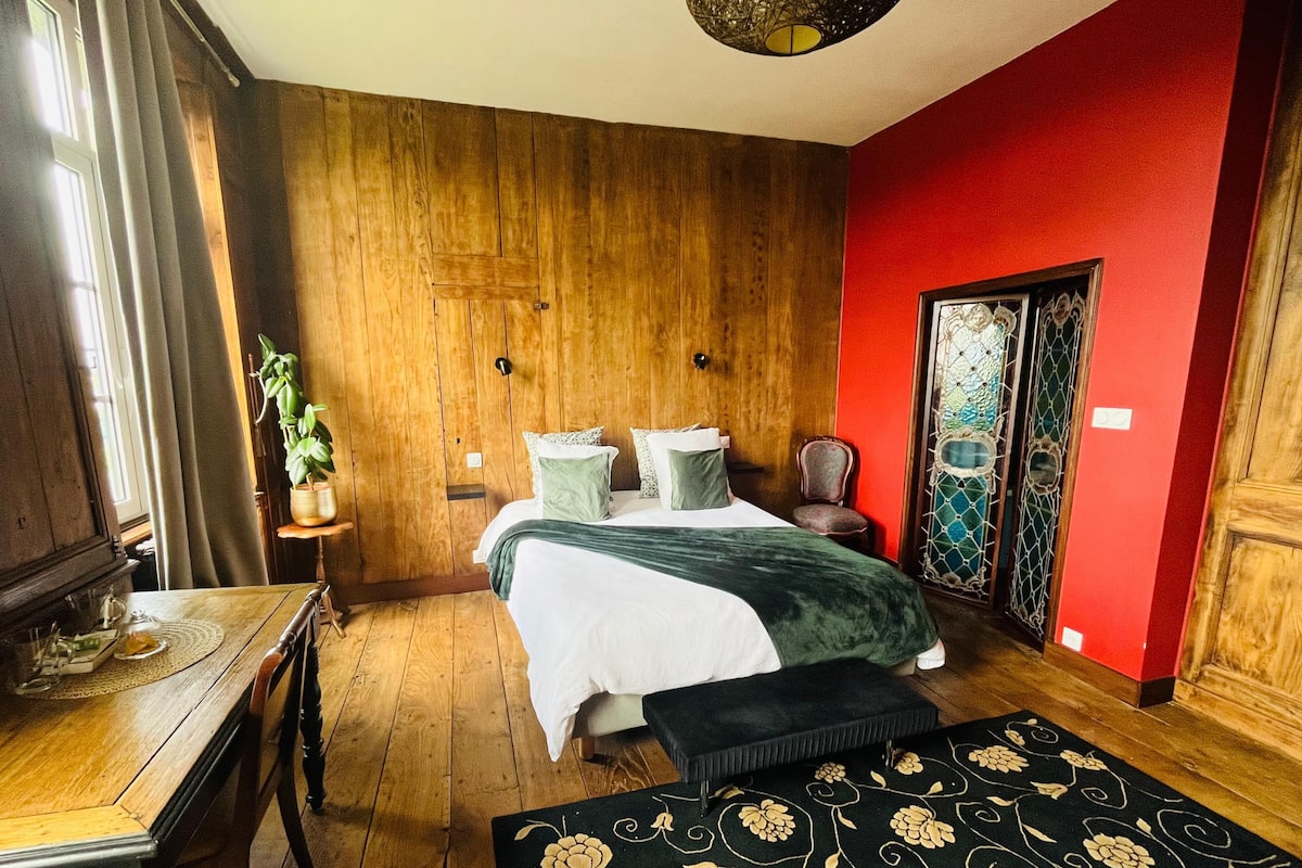Quarto com piso de madeira, cama com cobertor verde, uma cadeira, uma mesa pequena com uma planta e uma parede vermelha.