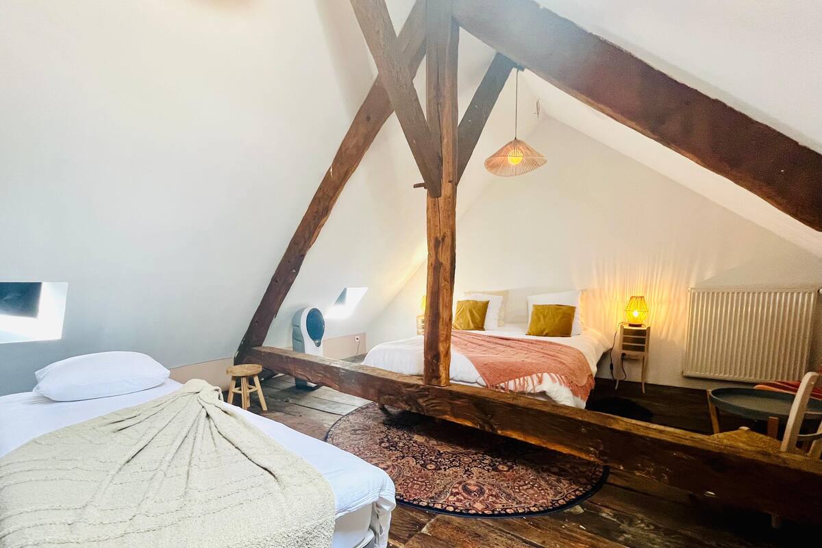 Quarto com teto inclinado, vigas de madeira, cama com cobertor branco e bege, um banquinho pequeno, tapete redondo estampado, uma mesa e uma cadeira.