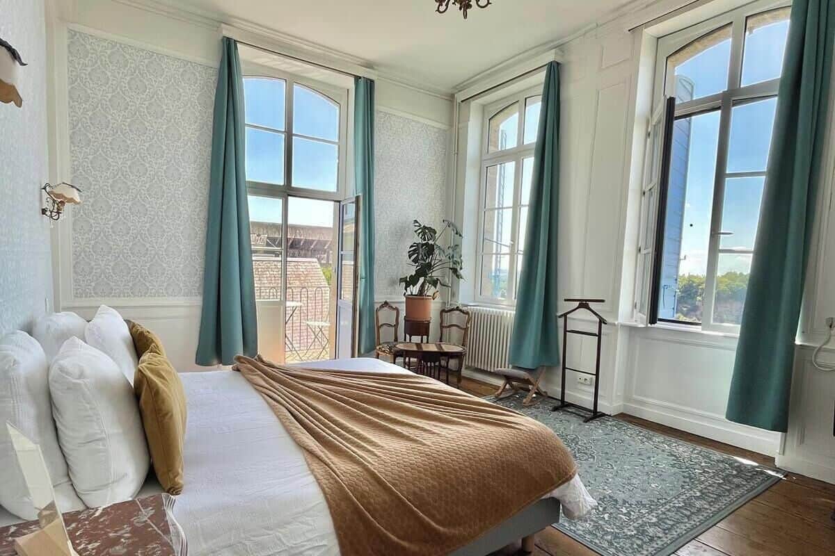 Quarto com cama, cômoda de madeira, pequena mesa, cadeira, planta em vaso e vista para o exterior por grandes janelas.