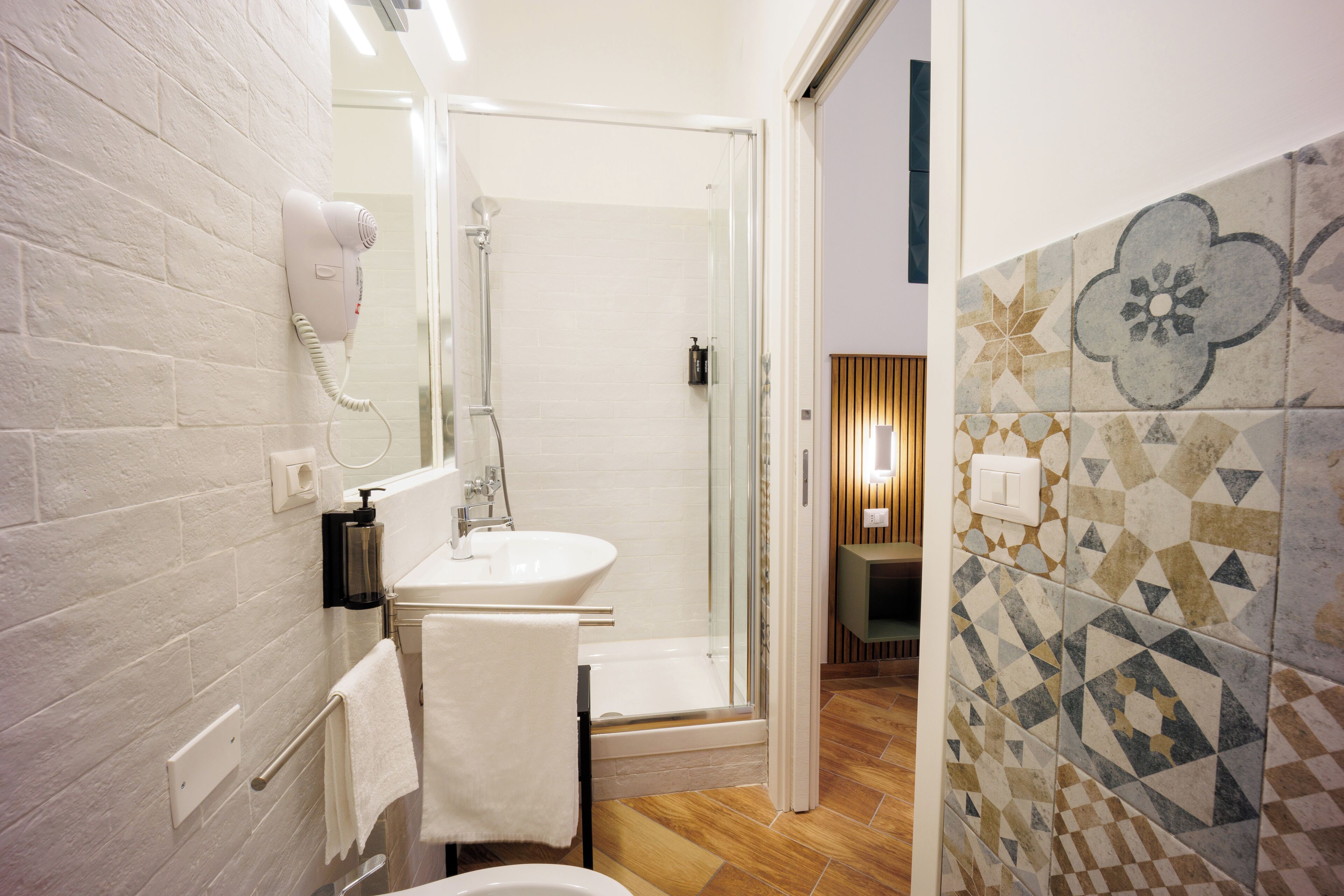 Um banheiro moderno com parede de tijolos brancos, box de chuveiro de vidro, pia branca e painel de azulejos estampados.