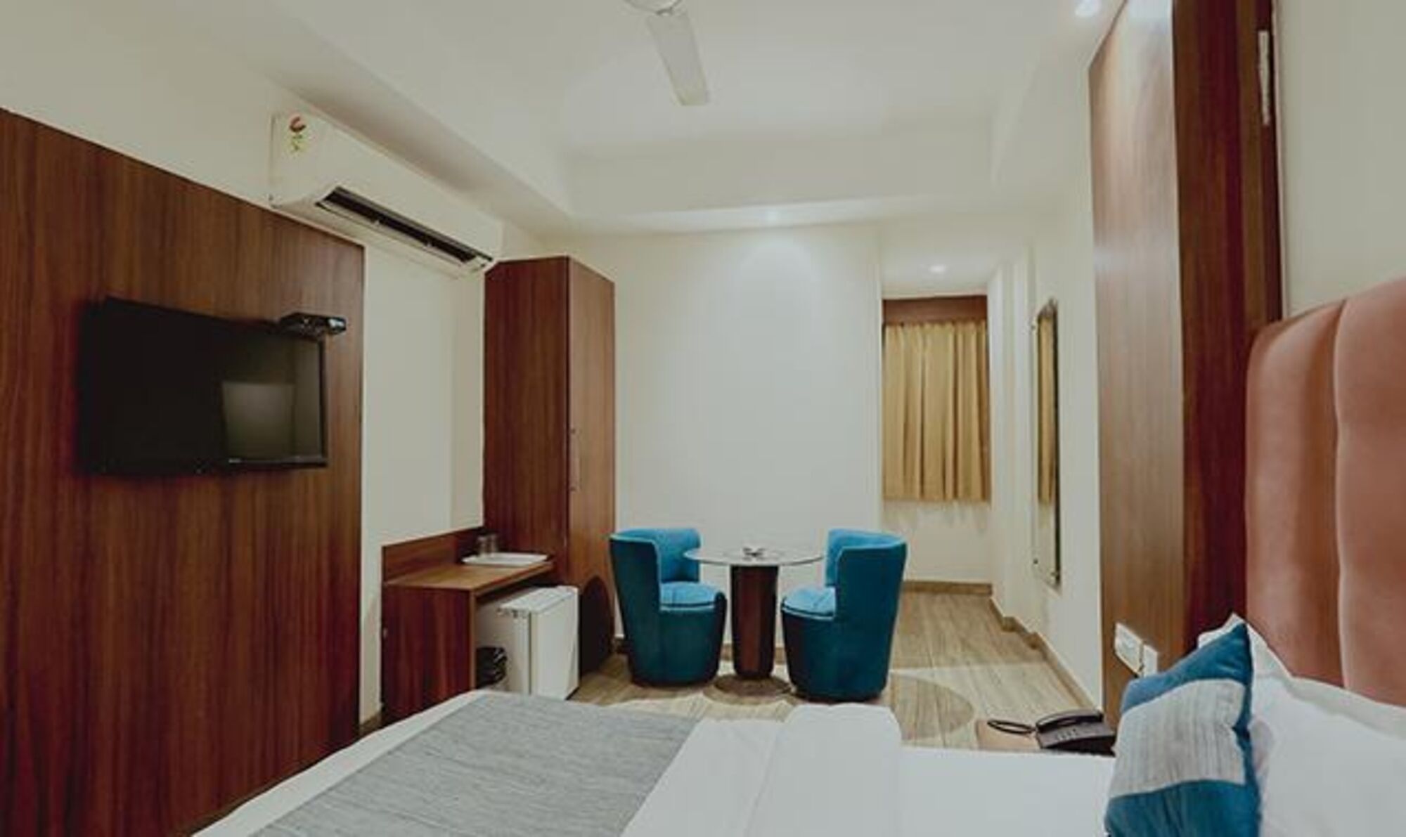 Quarto de hotel com cama, escrivaninha, duas cadeiras, televisão e janela com cortinas.