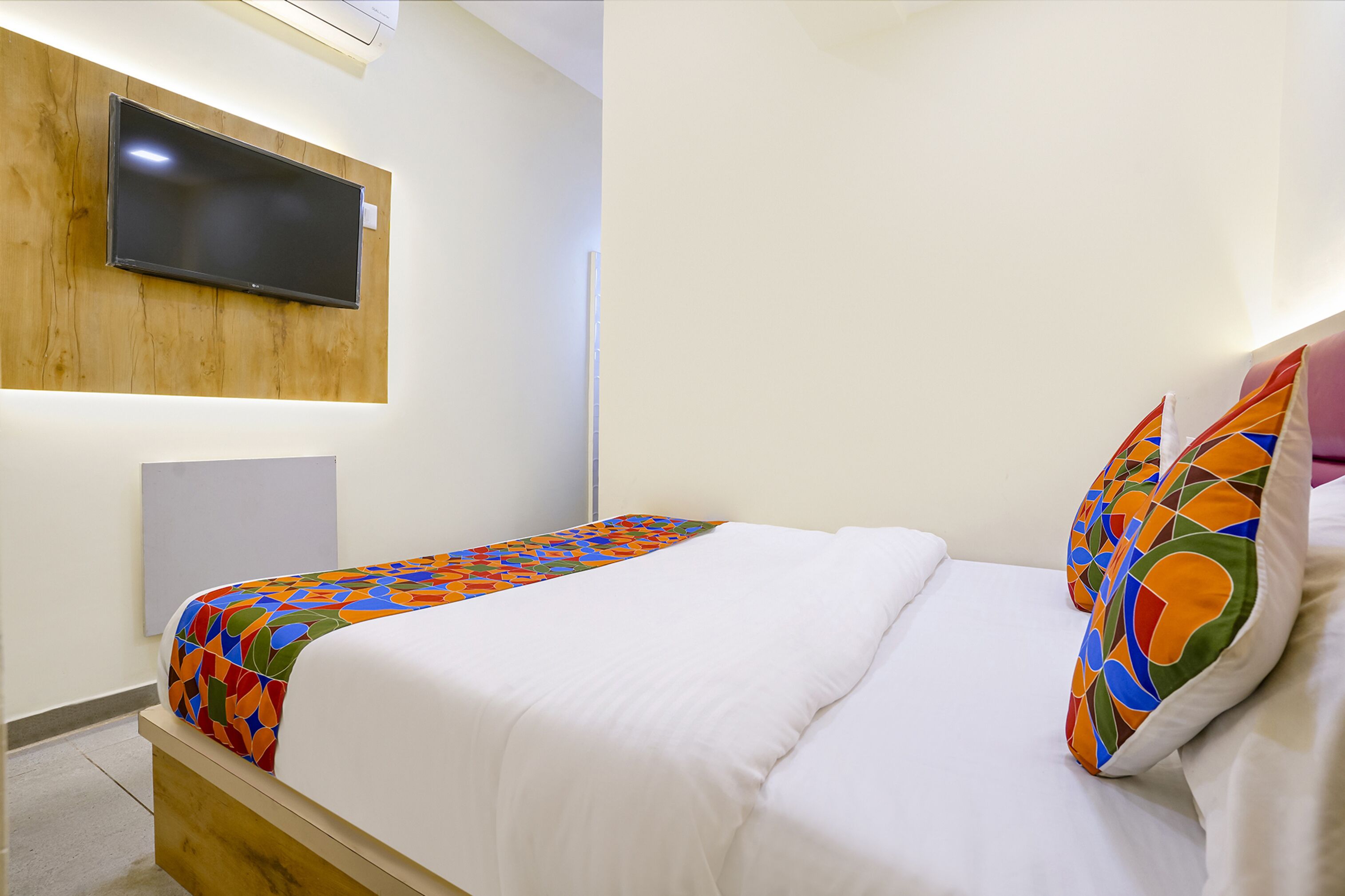 Uma cama de solteiro com colcha estampada em padrão geométrico colorido e travesseiros brancos, uma TV de tela plana montada na parede e cabeceira de madeira.