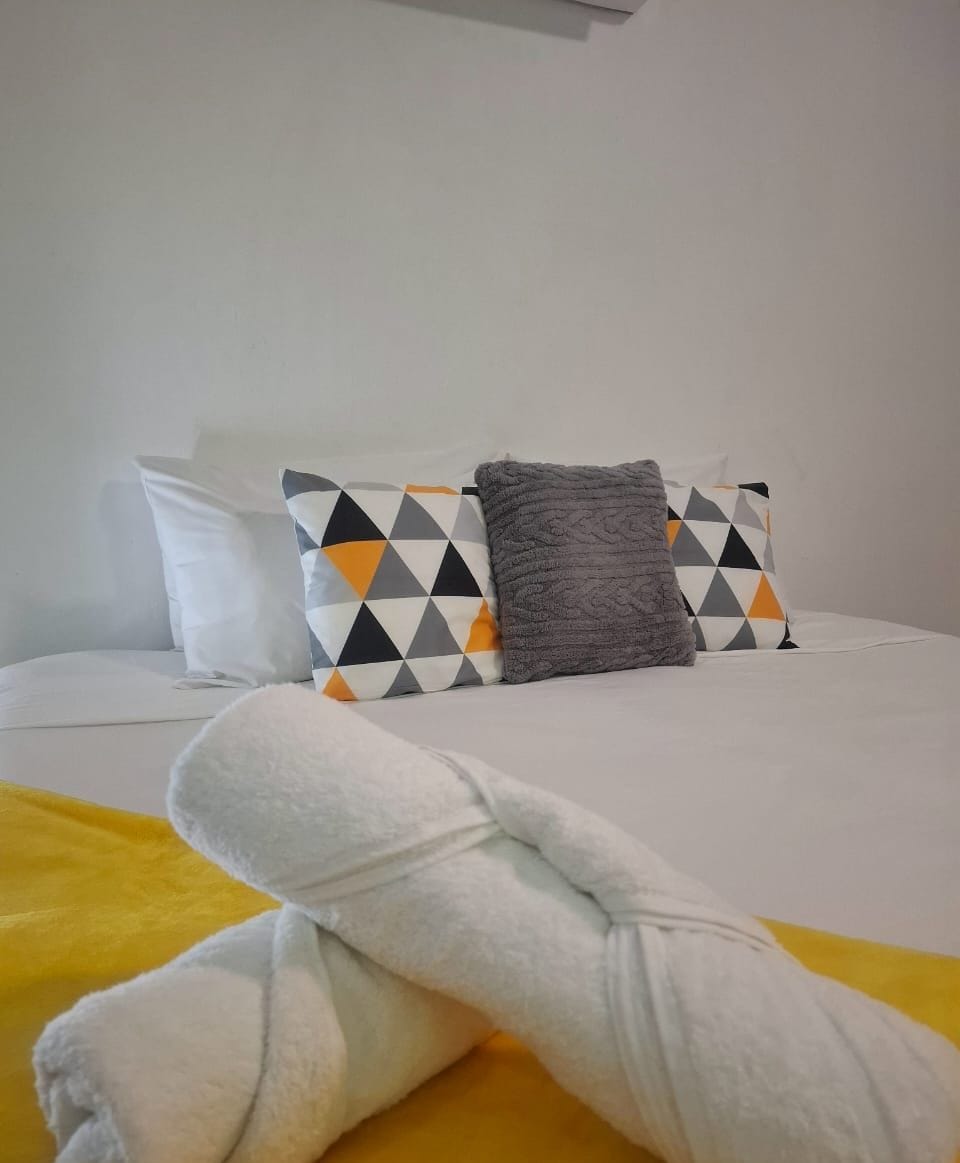 Cama com colchão branco, manta cinza e almofadas com padrões geométricos.