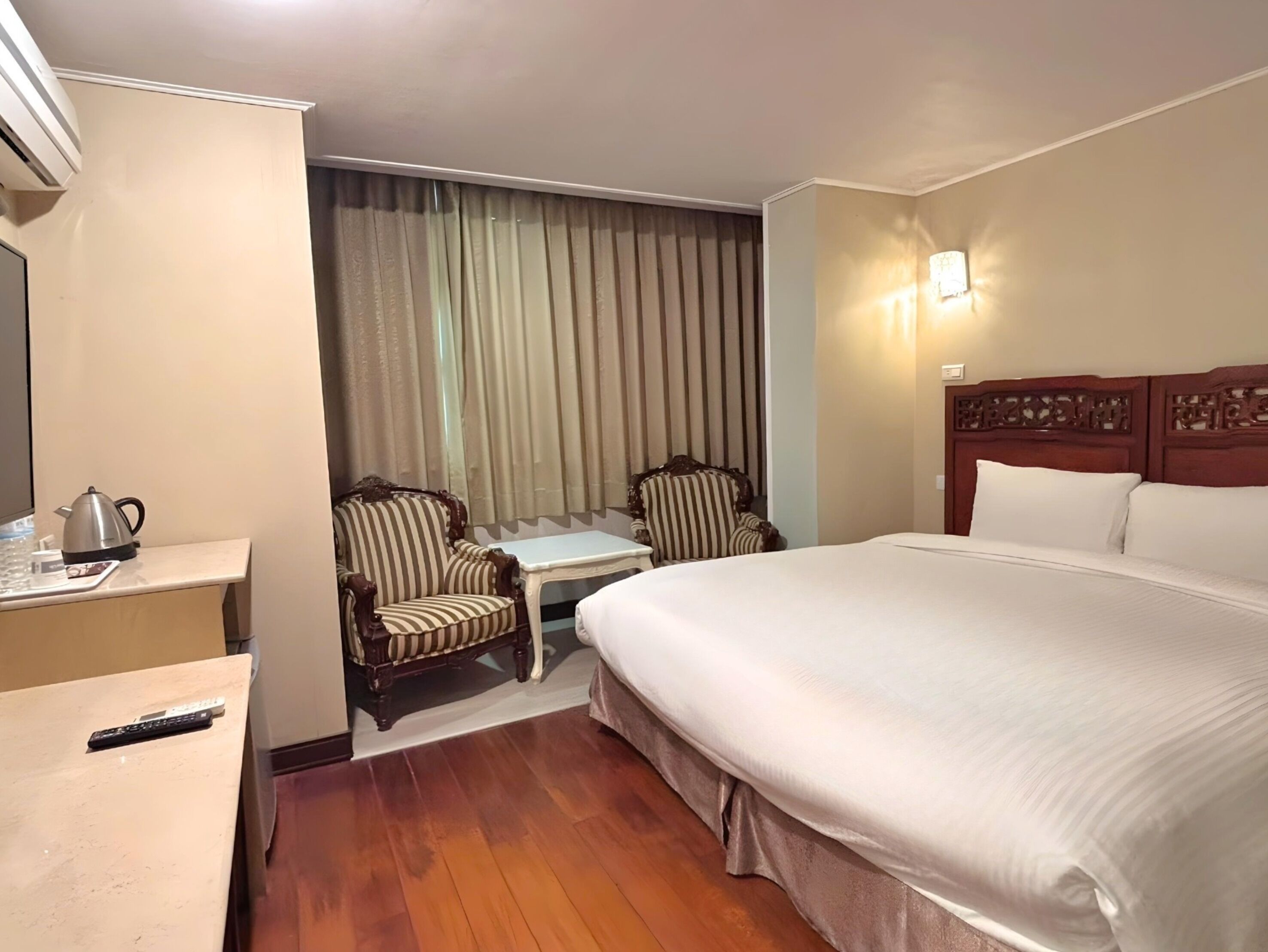 Quarto de hotel com uma cama grande, uma escrivaninha com cadeira, uma mesinha com chaleira e uma televisão.