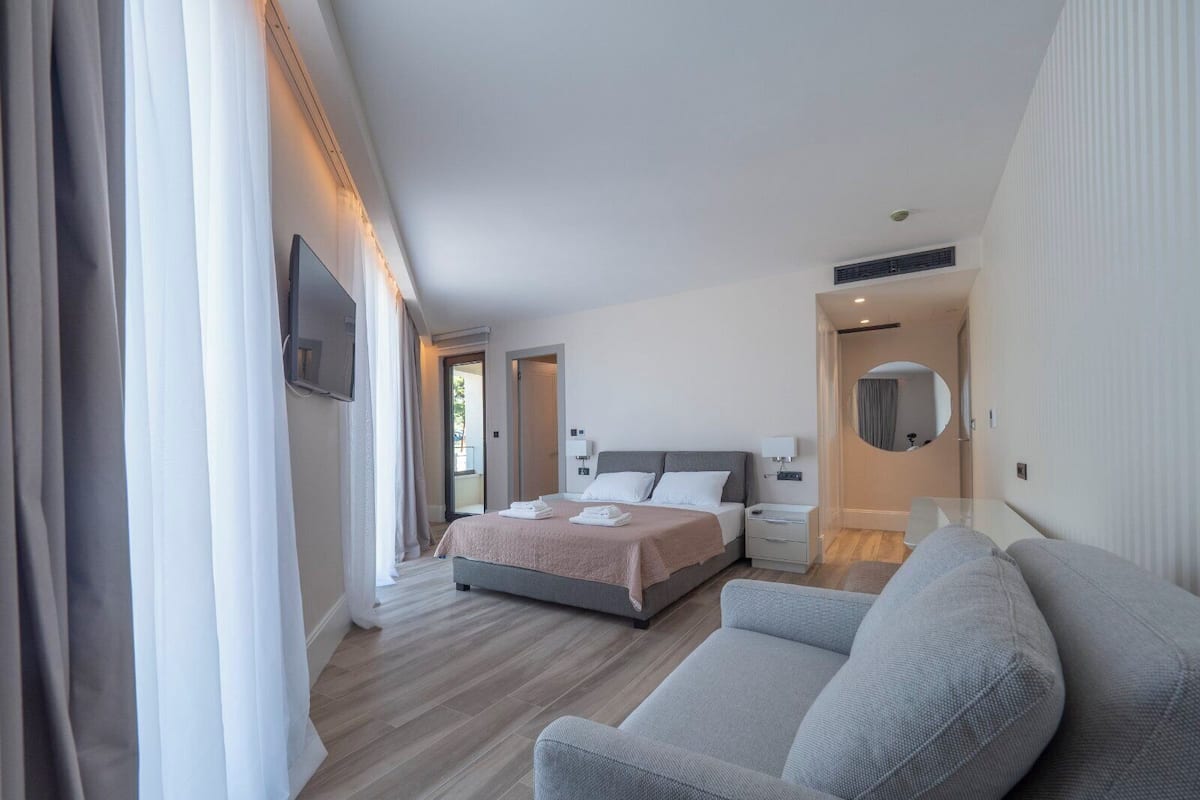 Quarto de hotel moderno com uma cama grande, um sofá, uma mesa de trabalho e um banheiro visível através de uma porta aberta.