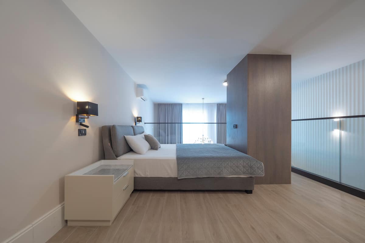Quarto moderno com cama grande, mesas de cabeceira, luz acoplada à parede e vista da cidade através de uma janela com cortinas.