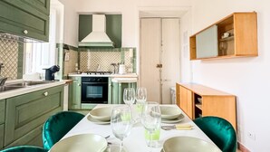 Uma área de refeições com uma mesa posta para quatro pessoas, cadeiras verdes e uma cozinha ao fundo.