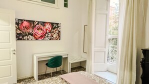 Quarto com cama, escrivaninha, cadeira, um grande quadro floral e vista para o exterior.