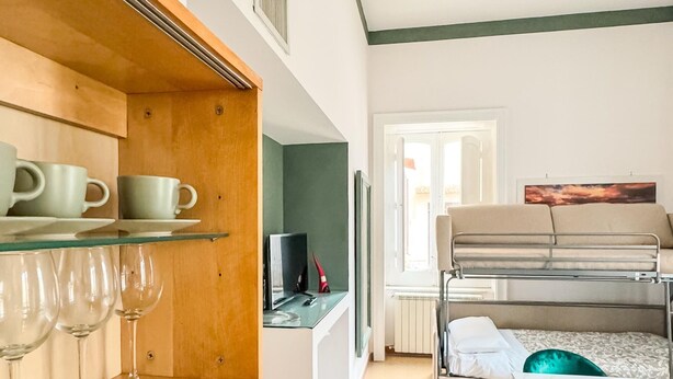 Quarto de hotel com prateleira de madeira com copos e taças, uma cama beliche com uma cadeira verde e um quadro na parede.