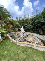 Uma fonte de pedra com uma estátua de Buda no centro, rodeada por vegetação exuberante e um céu límpido.