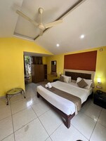 Quarto de hotel com cama, mesa de cabeceira, escrivaninha, cadeira, ventoinha no teto e janela com cortinas.