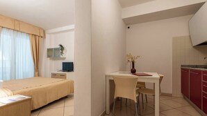 Quarto de hotel com cama, escrivaninha com cadeira, televisão, uma planta pequena e uma cozinha.