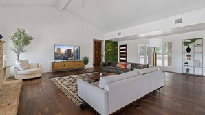 Uma sala de estar espaçosa com um sofá grande, uma mesa de centro e uma televisão. Há um armário de madeira e uma planta no lado esquerdo.