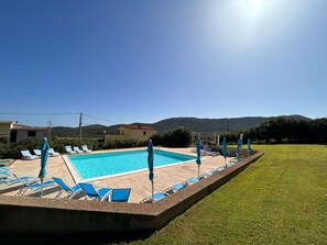 Piscina com espreguiçadeiras e guarda-sóis, rodeada por um gramado e colinas ao fundo.