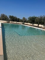 Piscina límpida com borda arenosa, rodeada por árvores e céu azul.