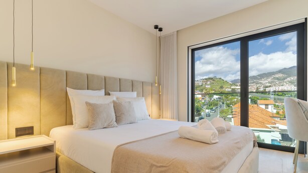 Quarto de hotel moderno com uma cama grande, mesa de cabeceira e vista para a cidade através de uma janela ampla.