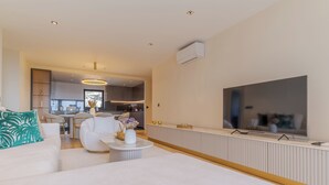Uma sala de estar moderna com uma grande TV de tela plana, um sofá, uma mesa de centro e uma área de jantar ao fundo.