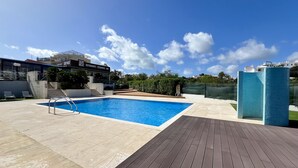 Uma piscina com deck de madeira, um restaurante chamado "Sexy Meat" ao fundo, e um céu azul límpido.