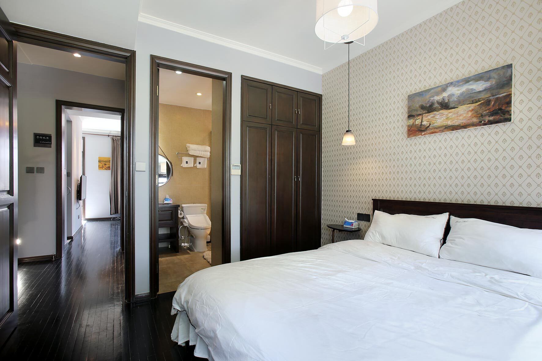 Quarto de hotel com uma cama grande, um guarda-roupa de madeira, casa de banho com sanita e lavatório, e um quadro na parede.