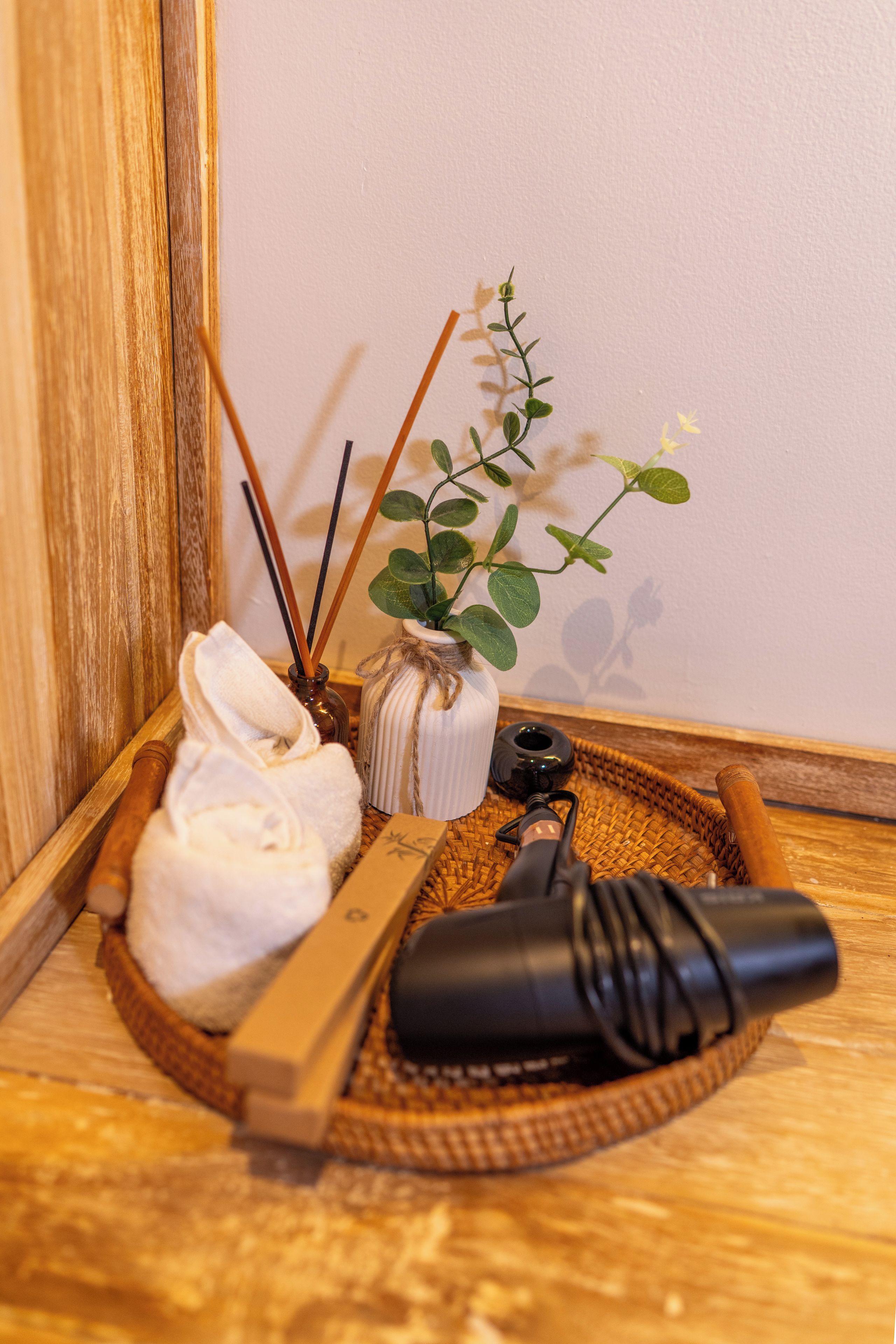 Canto de quarto com piso de madeira, uma cesta trançada contendo uma pistola de massagem preta, uma toalha branca, uma pequena planta em vaso e um frasco de óleo essencial.