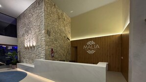 Uma área de recepção com parede de pedra, balcão de madeira e uma placa com a inscrição 'MALIA BEACH EXPERIENCE'.