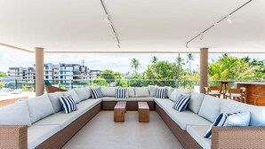 Uma área de estar moderna ao ar livre, com um amplo sofá modular e uma mesa de centro de madeira, com vista para um horizonte urbano e vegetação exuberante.