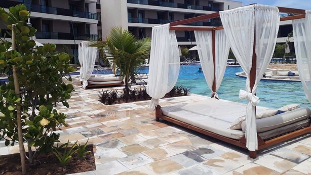 Área da piscina com espreguiçadeira, dossel e vista para um edifício com varandas.