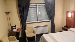 Quarto de hotel com cama, cadeira, mesinha, luminária e janela com cortinas.