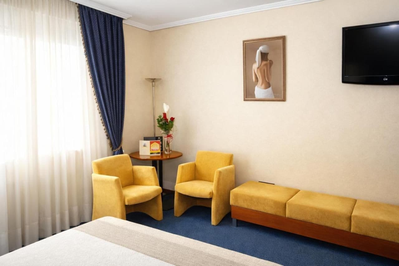 Quarto de hotel com uma cama, duas poltronas amarelas, uma mesa pequena com um vaso e uma caixa, um sofá, um quadro e uma televisão.
