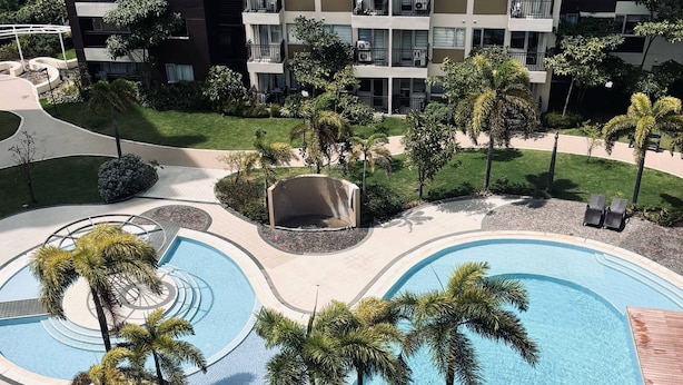 Um edifício residencial com vários níveis, varandas, área de piscina ajardinada com palmeiras e um pequeno jardim.