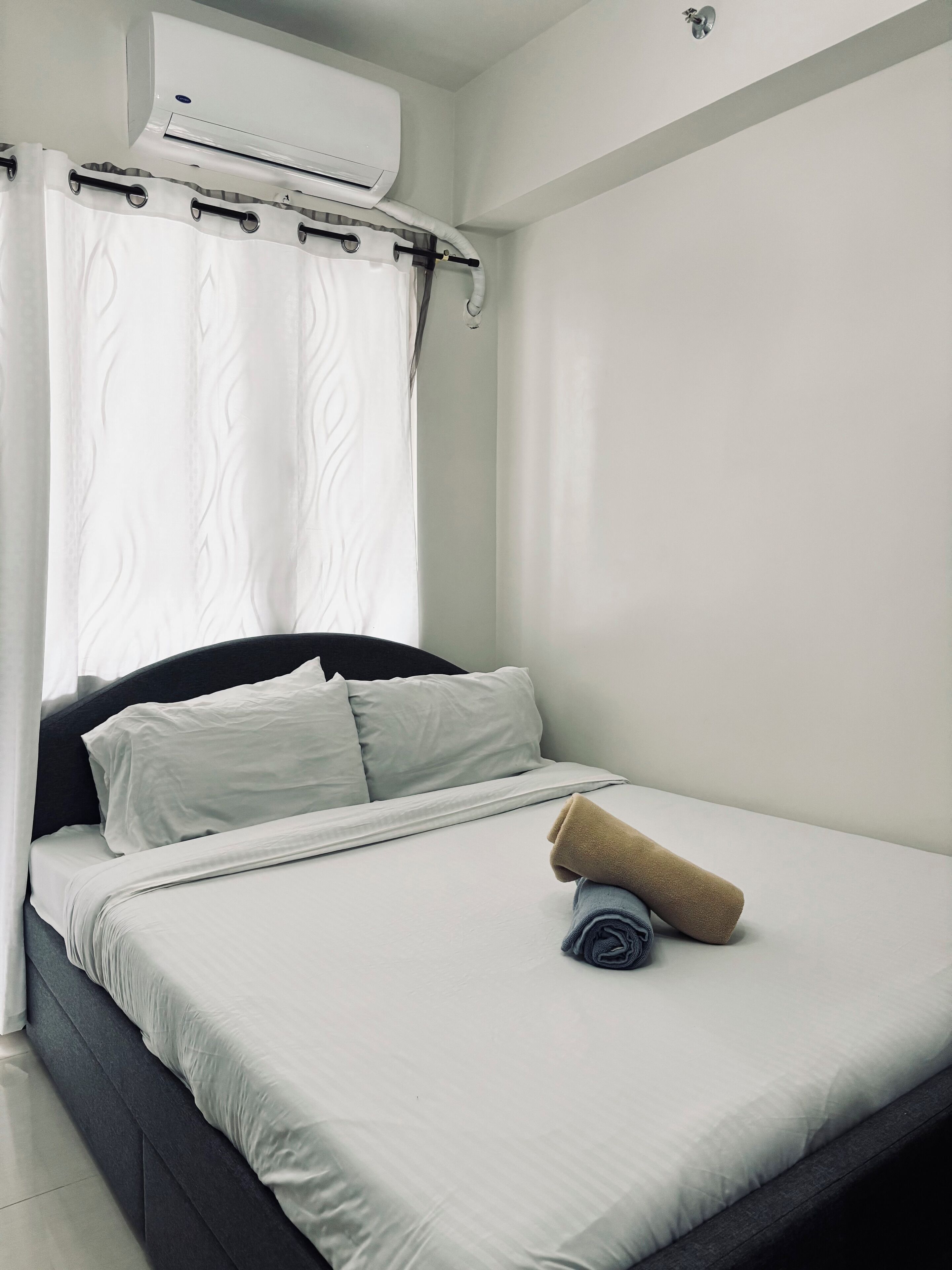 Cama bem arrumada com lençóis brancos, toalha enrolada e cabeceira escura.
