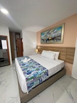 Quarto de hotel com uma cama, um quadro de barcos na parede, uma mesa de cabeceira e um banheiro visível ao fundo.