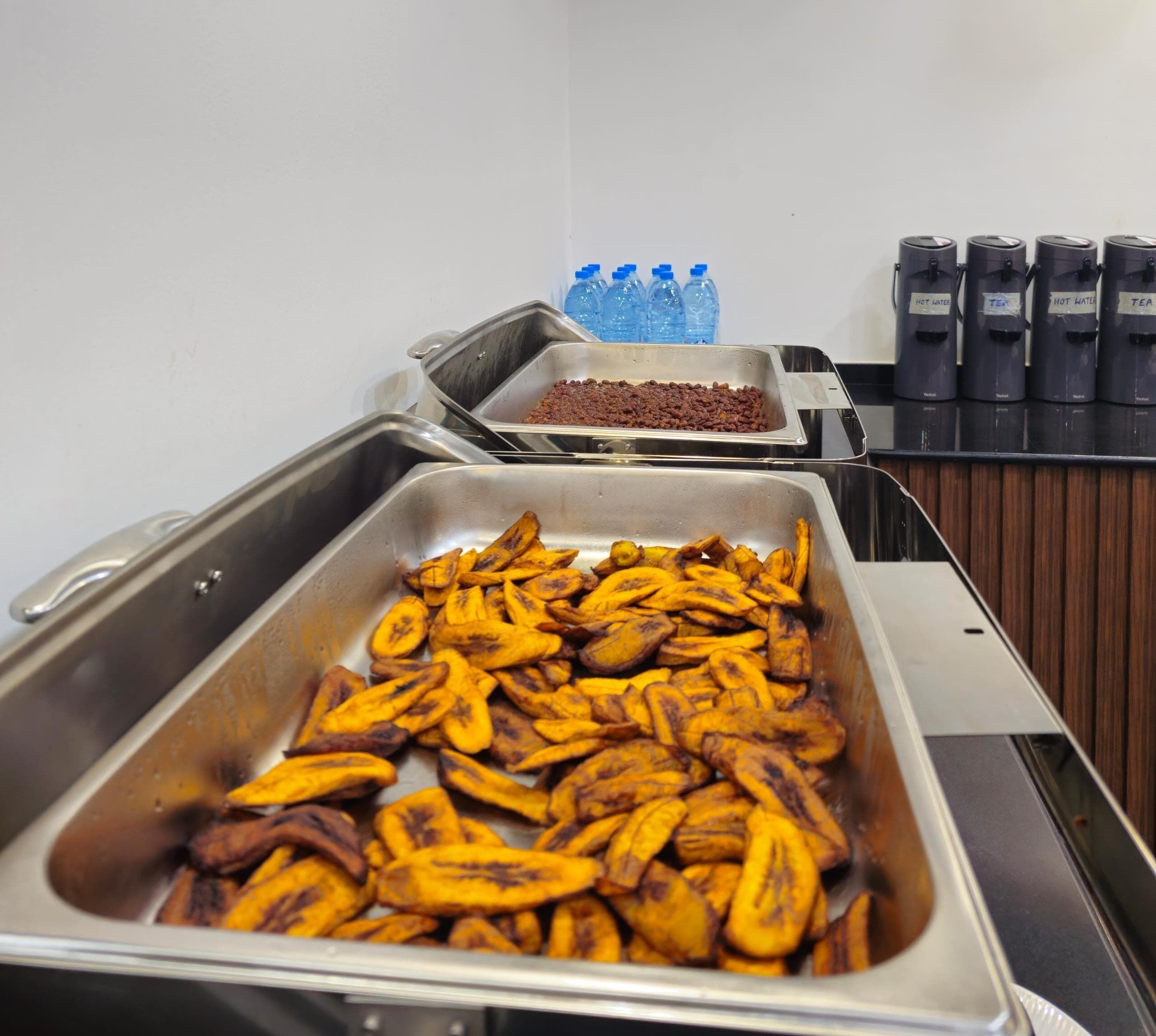 Buffet com bandejas de comida, incluindo bananas e creme de chocolate. Há também garrafas de água e dispensadores de chá.