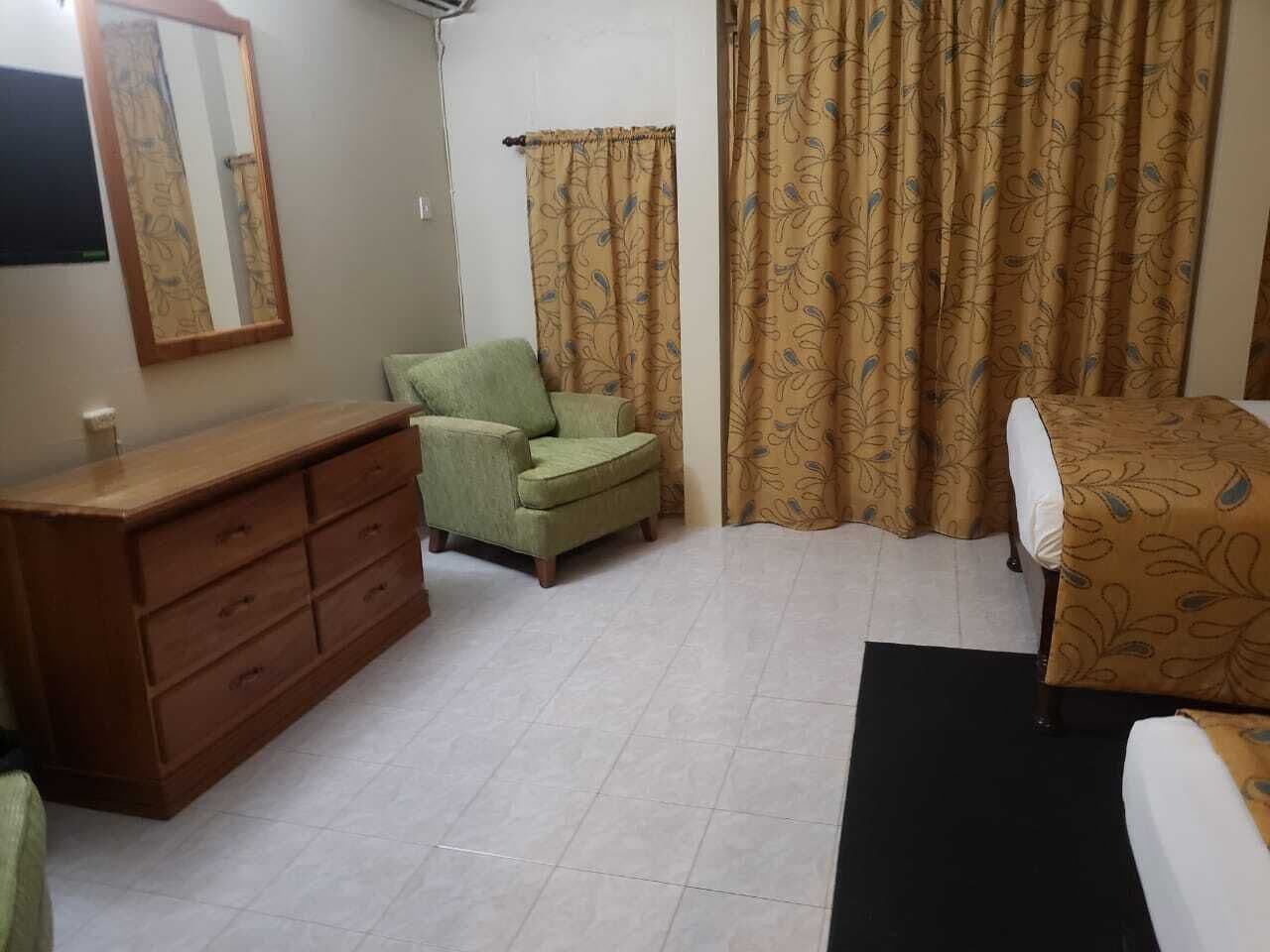 Um quarto com uma cama, uma poltrona verde, um aparador de madeira e cortinas estampadas.