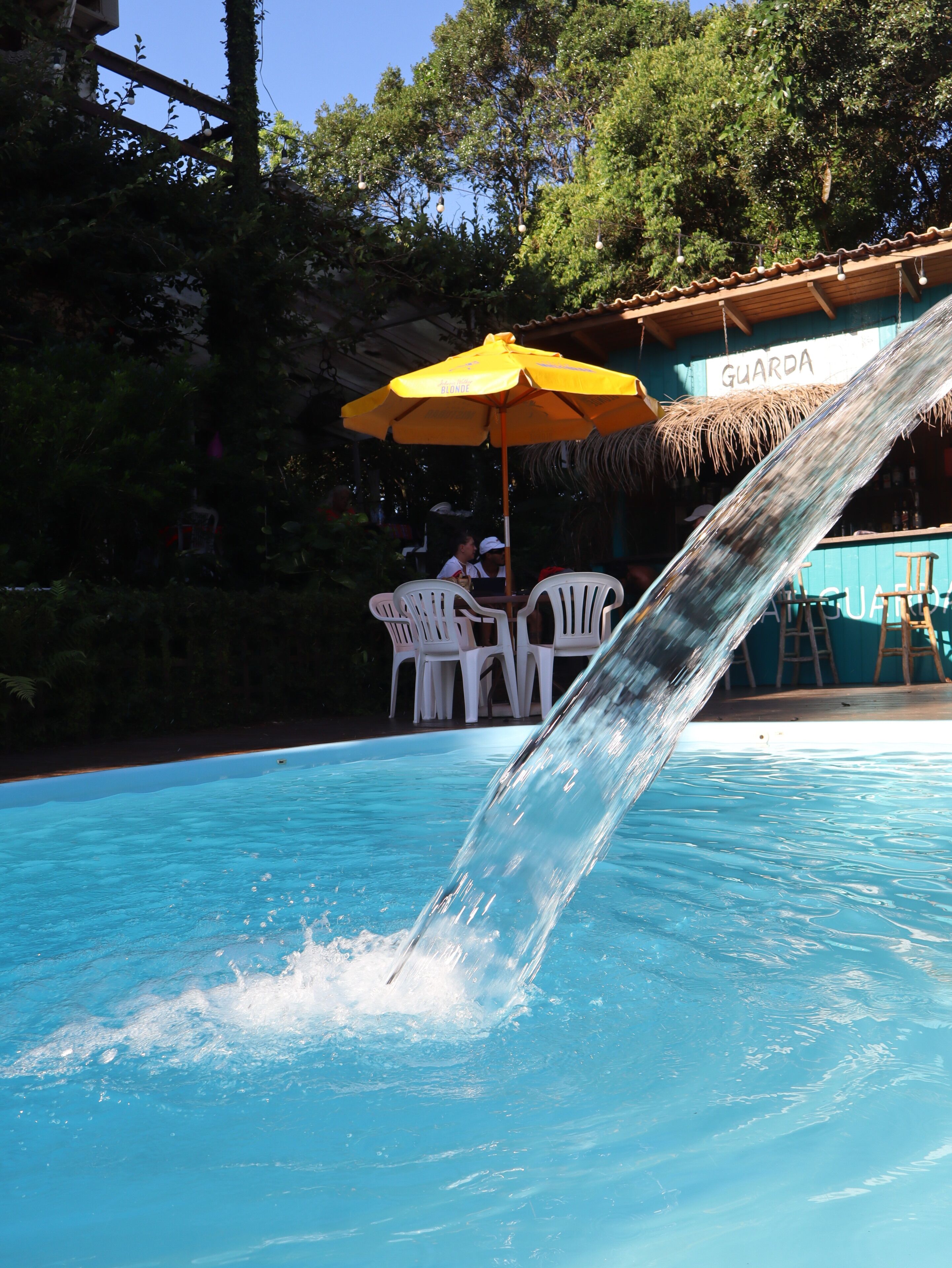 Piscina com escorregador, cadeiras de plástico branco e um guarda-sol amarelo.