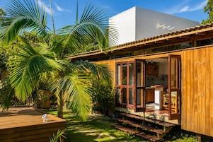 Um deck de madeira com uma palmeira, um prédio branco e uma pérgula de madeira com portas de vidro.