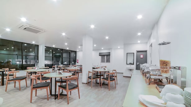 Uma área de refeições moderna, com mesas e cadeiras redondas, um balcão comprido com pratos brancos e uma grande janela com vista.