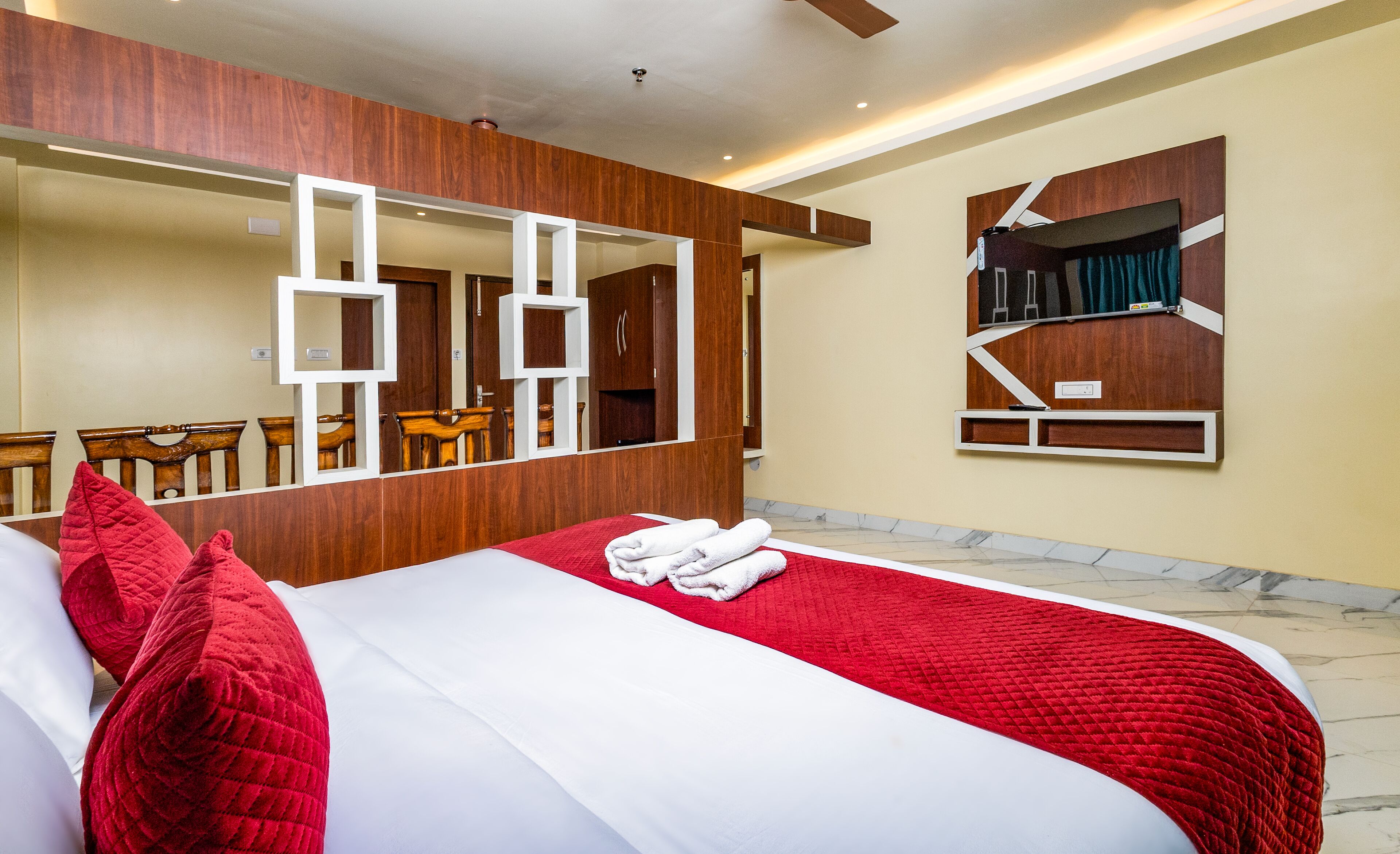 Quarto de hotel com uma cama grande, almofadas vermelhas, manta vermelha, cabeceira de madeira, um espelho grande e uma televisão fixada na parede.