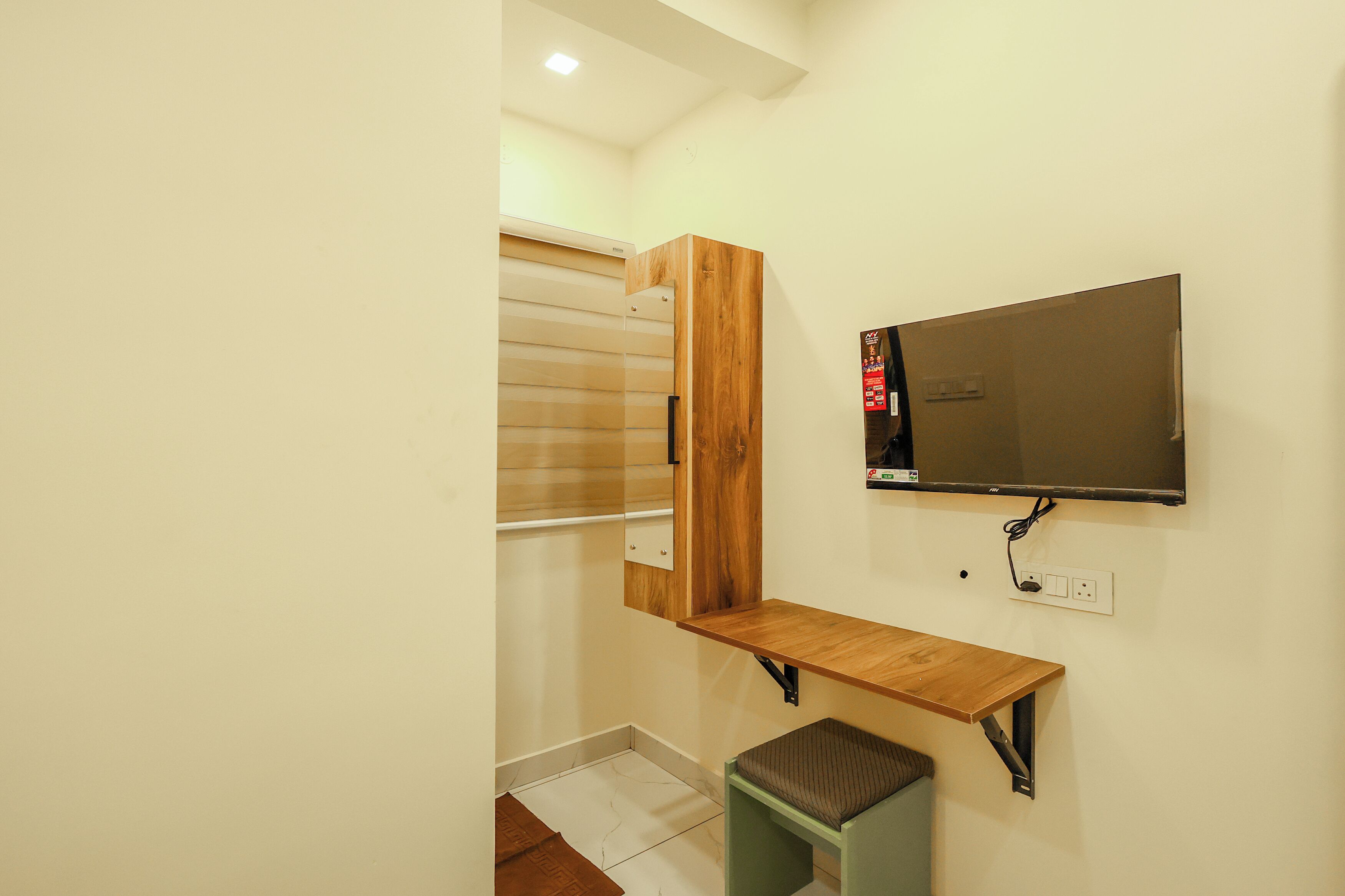 Quarto pequeno e moderno, com um banco de madeira, televisão na parede e um banquinho.