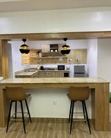 Cozinha moderna com ilha central, dois bancos de bar, armários de madeira e iluminação pendente.