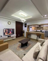 Uma sala de estar moderna com uma televisão de tela plana, uma mesa de centro escura e um sofá. Há uma área de cozinha com um balcão e banquetas.