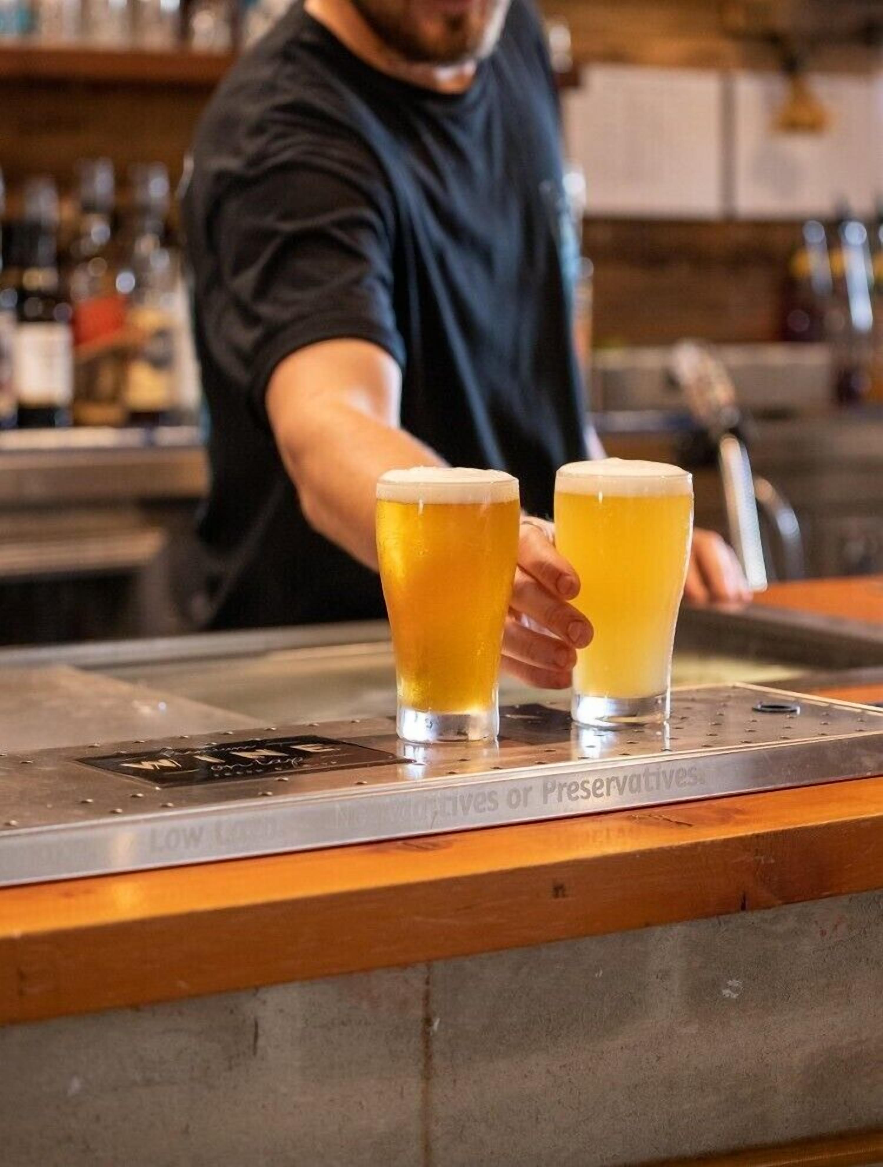 Dois copos de cerveja em um balcão de bar com superfície metálica e borda de madeira.