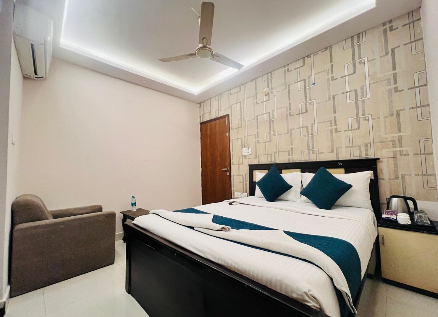 Quarto de hotel com cama, cadeira, mesa de cabeceira, garrafa de água e ventoinha no teto.
